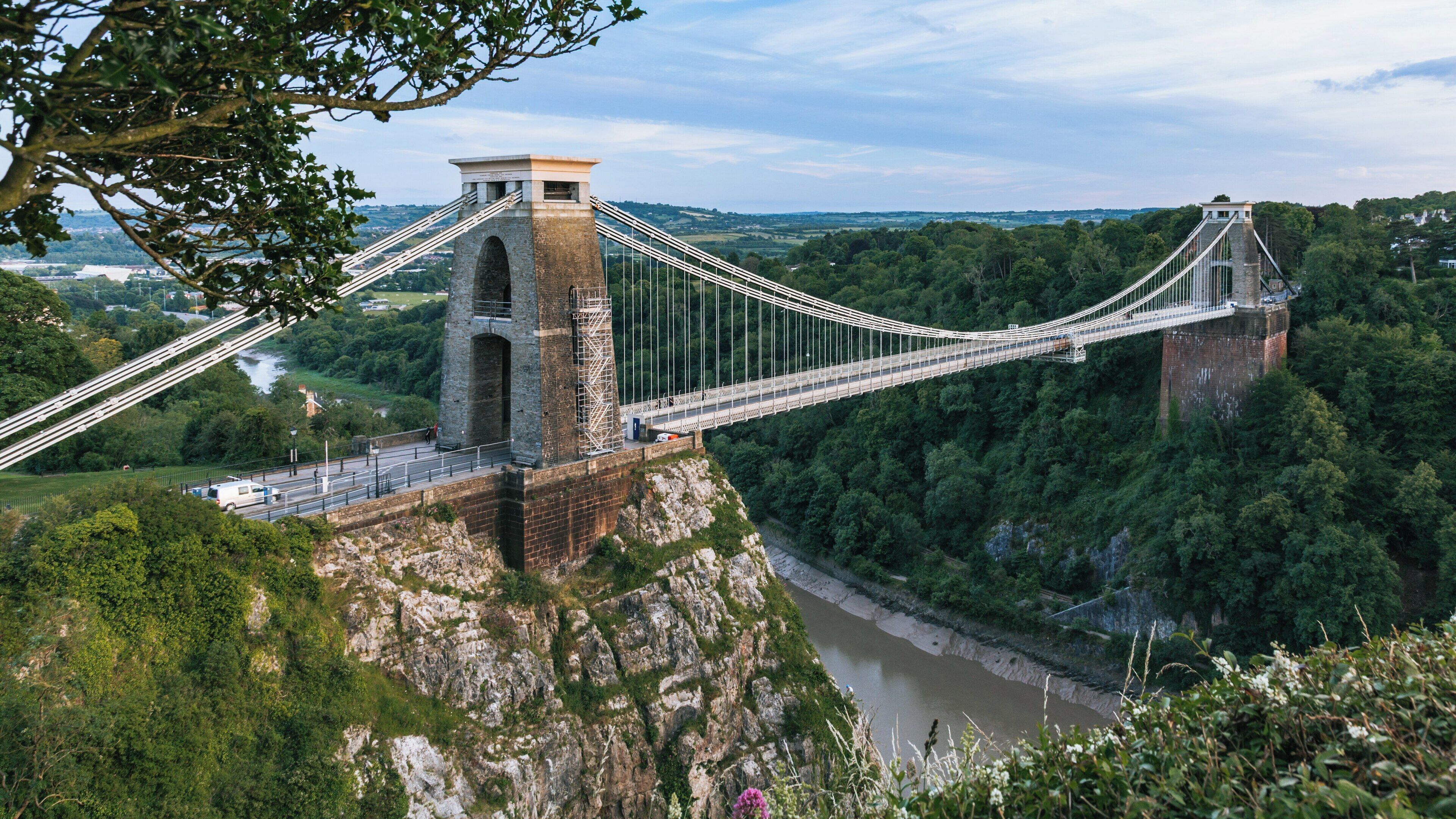 Uma ponte suspensa sobre um rio, cercada por vegetação exuberante e penhascos rochosos.
