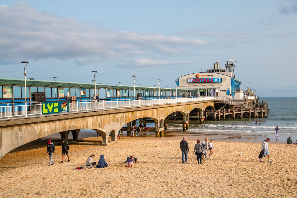 Um cais com um restaurante chamado "JACKTAILS AT" e uma praia onde pessoas caminham e crianças brincam.