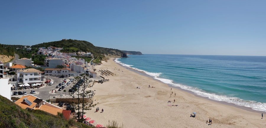 Uma vila costeira com uma praia arenosa, mar azul cristalino e edifícios numa encosta.