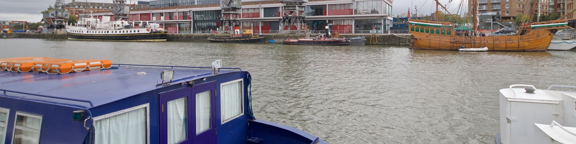 Um porto com um barco azul atracado em primeiro plano, um grande estaleiro com guindastes ao fundo, e um barco de madeira tradicional amarrado à direita.