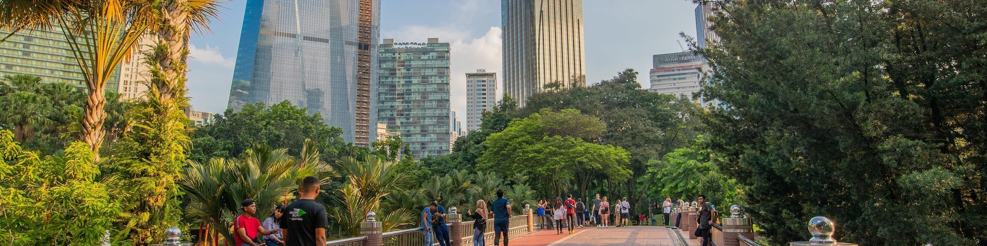 Uma ponte para pedestres com um caminho vermelho, rodeada por vegetação exuberante e edifícios altos ao fundo.