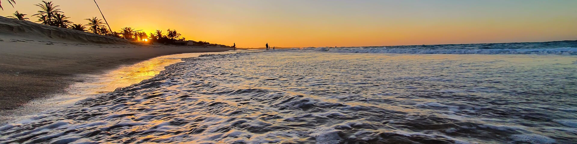 Praia ao pôr do sol com ondas, palmeiras e céu claro.