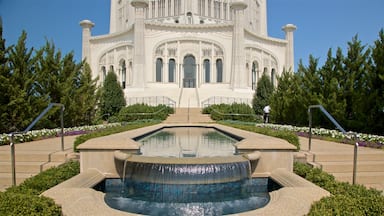Um edifício branco e imponente com arquitetura elaborada, uma fonte central e um jardim simétrico com árvores e flores.