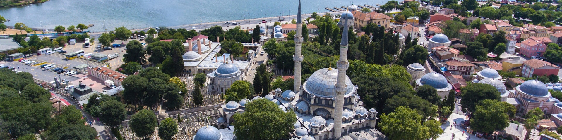 Paisagem urbana com uma mesquita em destaque, com várias cúpulas e um minarete alto, rodeada por árvores verdes e exuberantes e um rio.