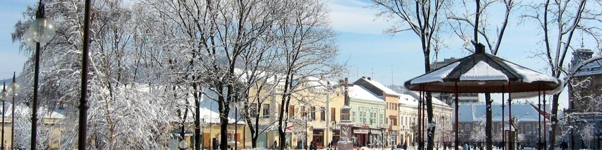Um parque coberto de neve com um coreto, árvores sem folhas e edifícios ao fundo.