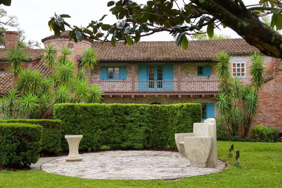 Uma casa tradicional de tijolos com telhado de telhas, cercada por sebes bem cuidadas e plantas tropicais.