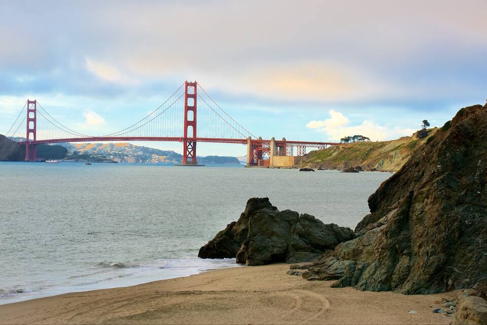 A Ponte Golden Gate, uma grande ponte suspensa, atravessa um corpo de água com um litoral rochoso e colinas ao fundo.