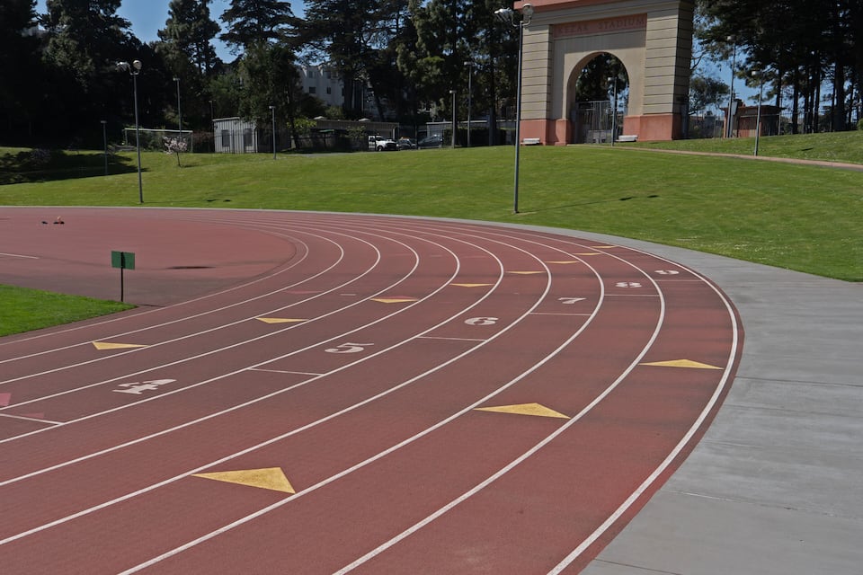 Pista de atletismo com raias, uma placa verde e um arco com a inscrição 'STADIUM' ao fundo.