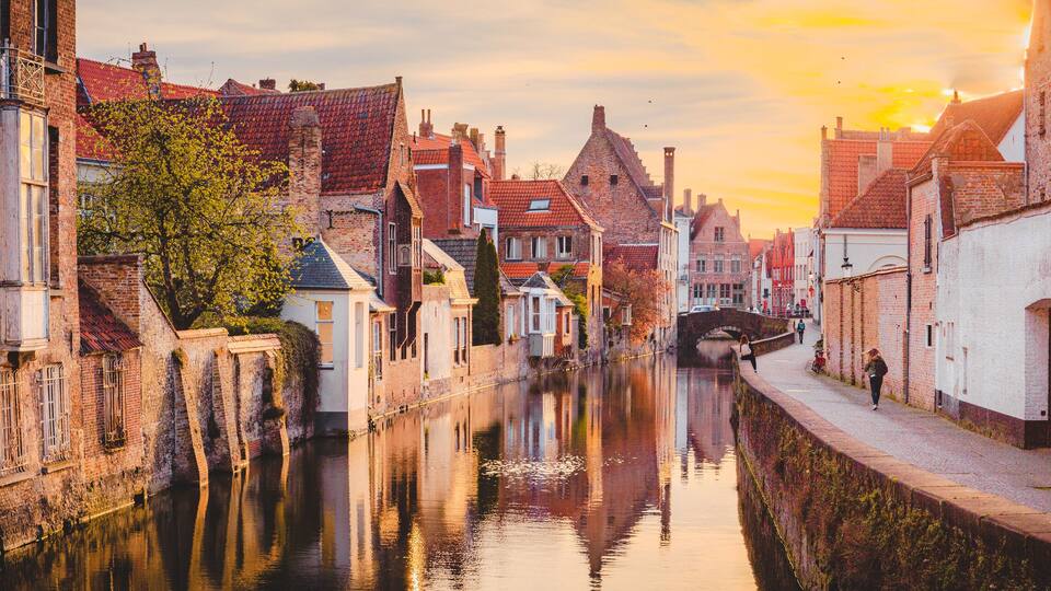 Um canal com edifícios de tijolo em ambas as margens, uma ponte ao longe e um céu ao pôr do sol.
