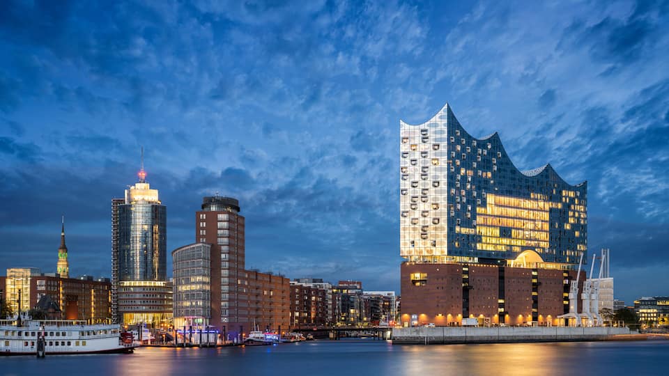 Horizonte urbano com um edifício moderno marcante, uma torre histórica e um calçadão tranquilo.