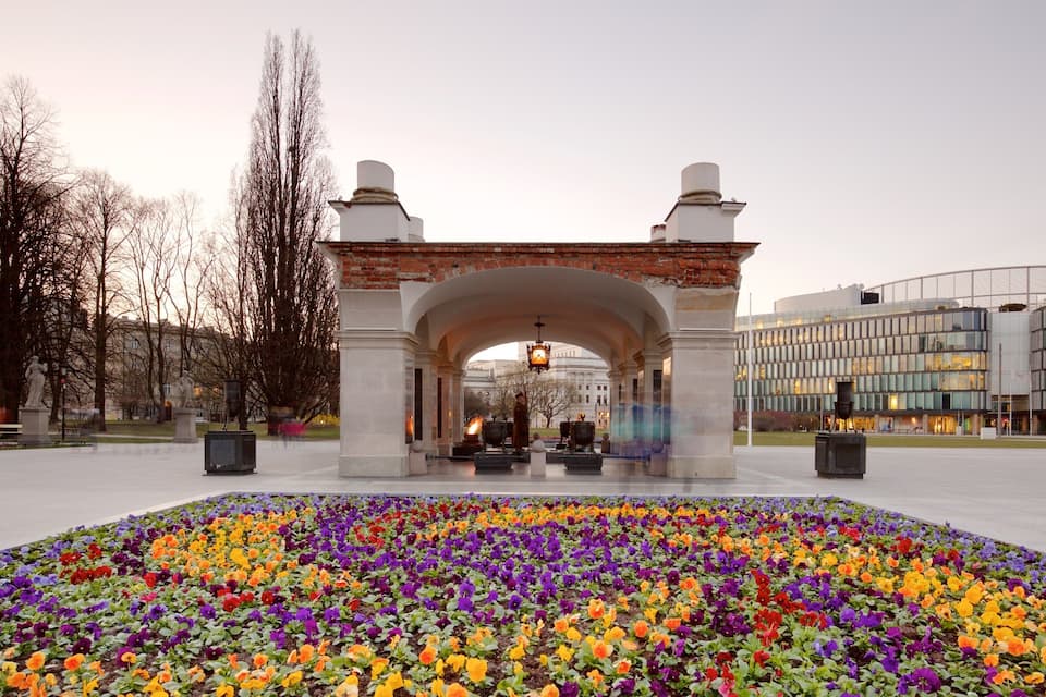 Um jardim de flores com um arco central e um edifício moderno ao fundo.