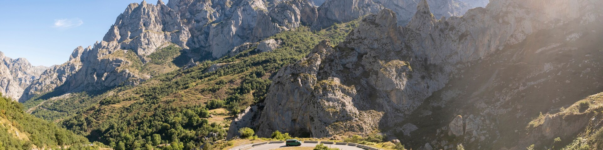 Uma estrada sinuosa na montanha com um carro verde, rodeada por penhascos rochosos e vegetação.