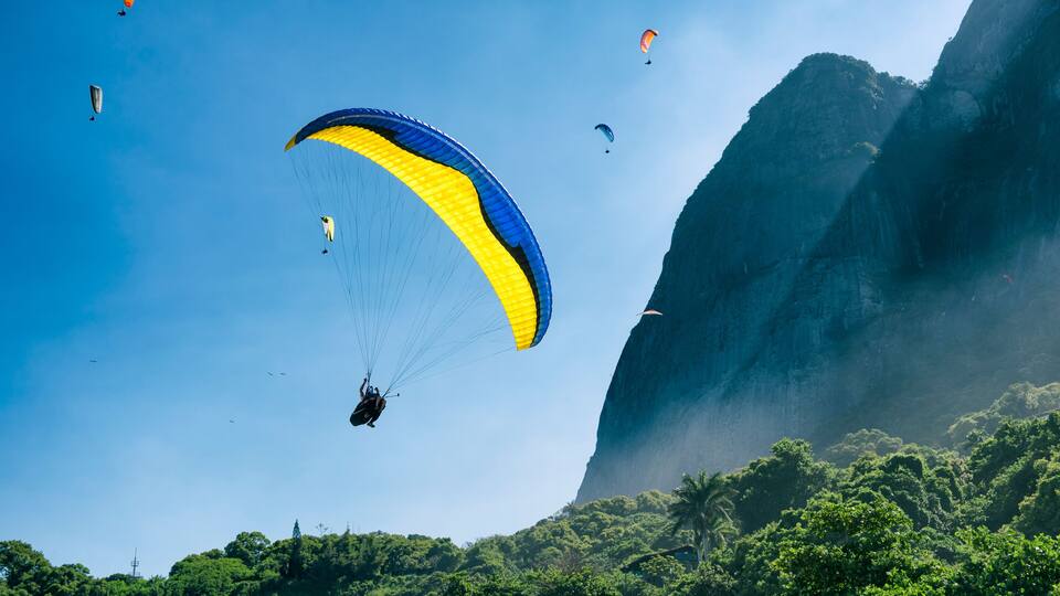 Um paraquedista planando sobre uma paisagem montanhosa, com um paraquedas azul e amarelo.