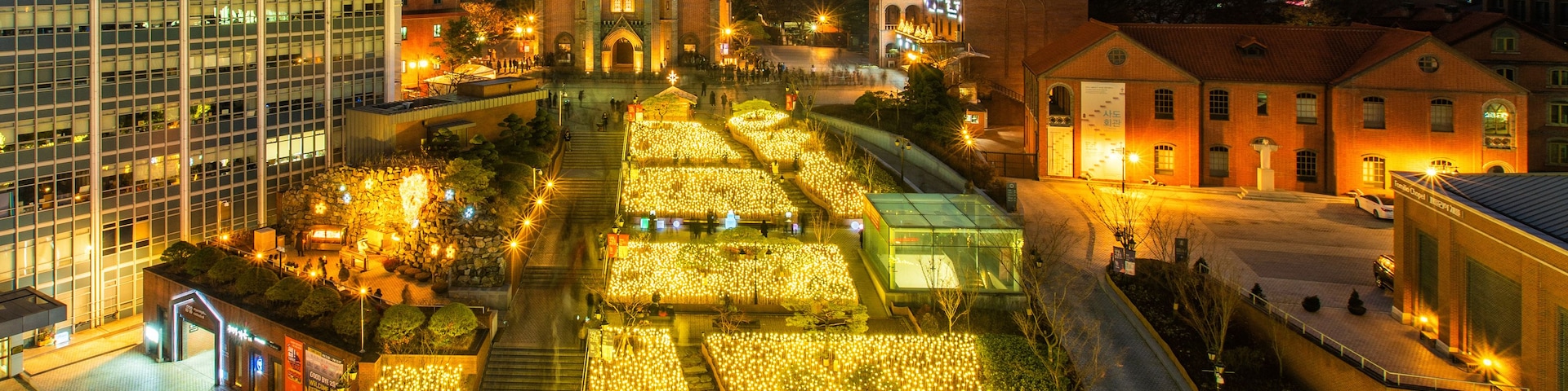 Paisagem urbana noturna com luzes de Natal iluminadas, uma igreja imponente com uma torre e edifícios modernos.