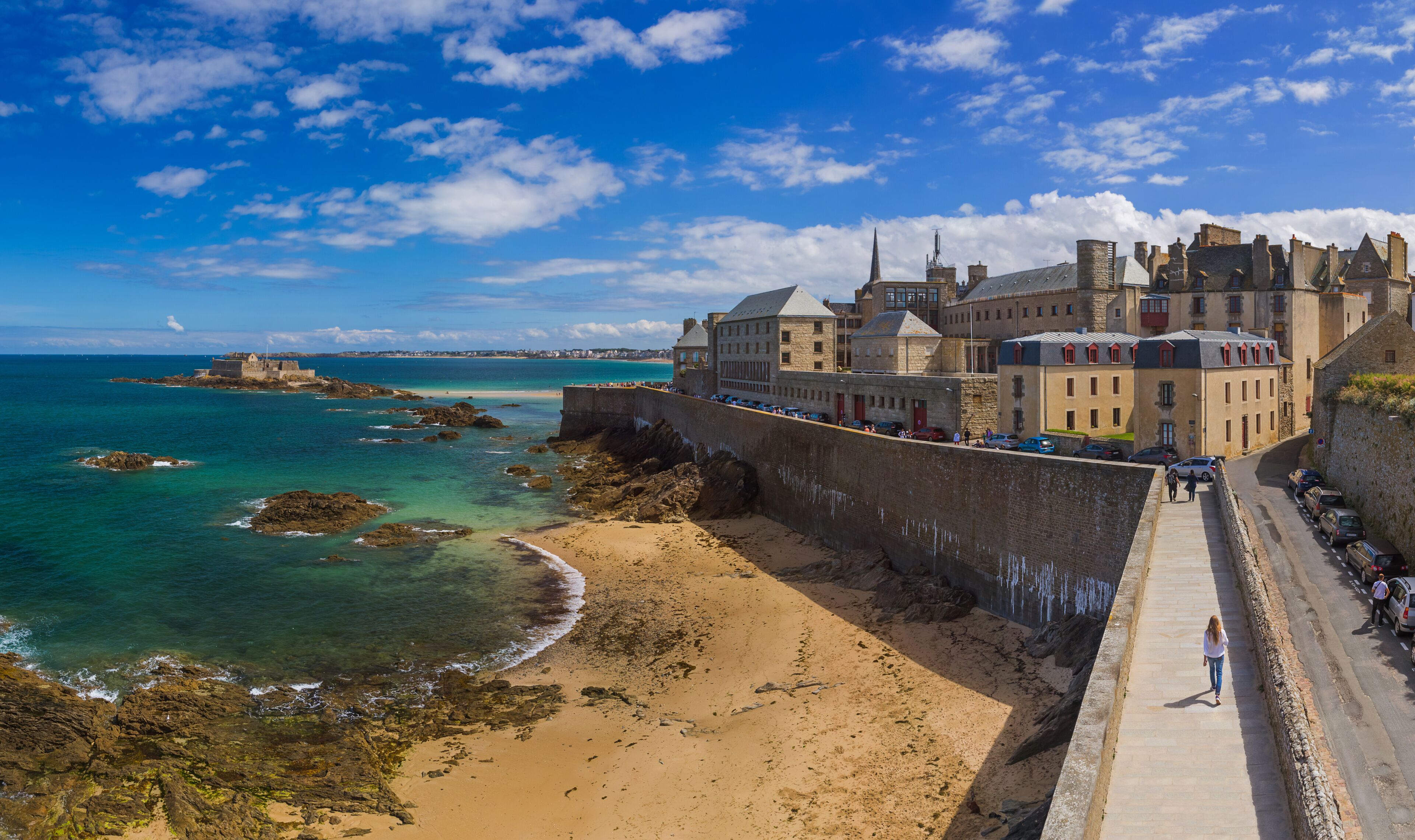 Uma vila costeira com um castelo imponente sobre o mar, uma praia de areia e um céu azul límpido.