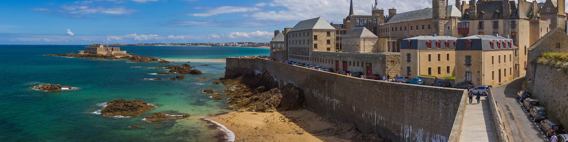 Uma vila costeira com um castelo imponente sobre o mar, uma praia de areia e um céu azul límpido.