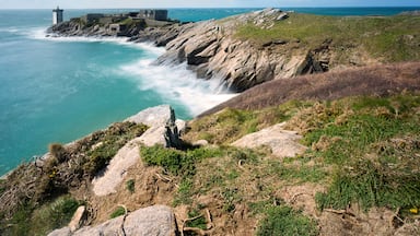 Paisagem costeira com falésias rochosas, um farol e uma estrutura fortificada à beira-mar.