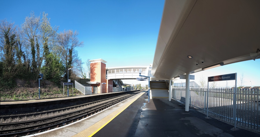 Plataforma de estação de trem com trilhos, área de espera coberta e uma ponte ao fundo.