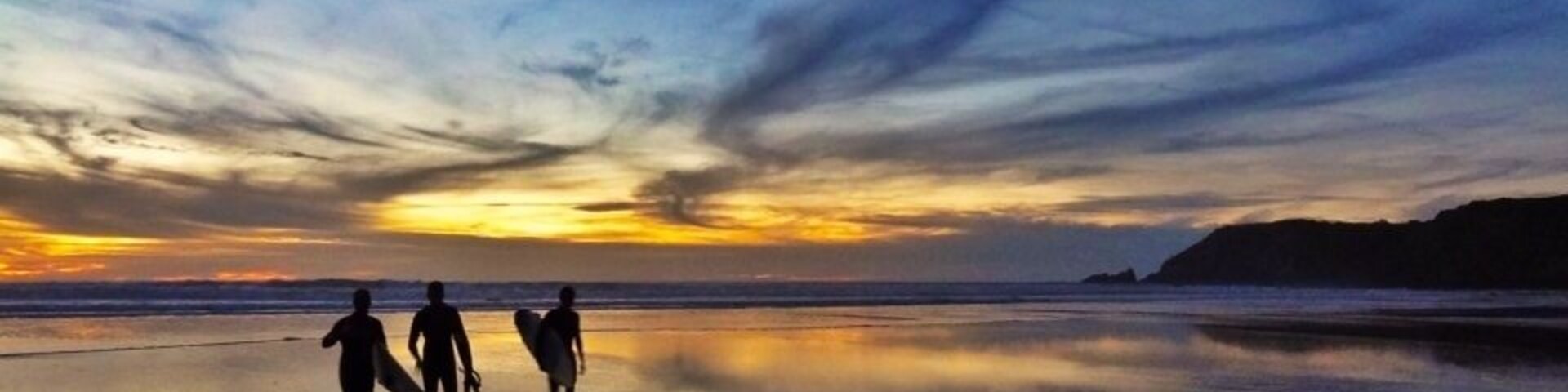 Praia ao pôr do sol, com silhuetas de pessoas caminhando e carregando pranchas de surf.