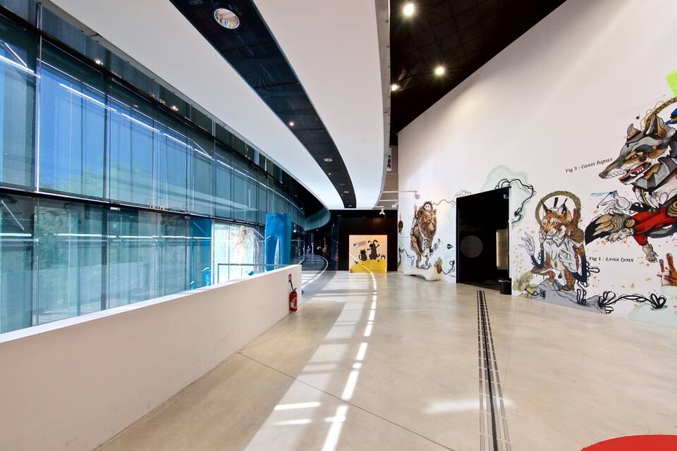 Interior de um edifício moderno com paredes de vidro, piso ladrilhado e murais coloridos nas paredes.
