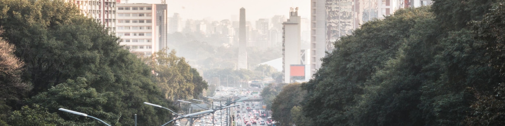 Uma autoestrada movimentada com múltiplas faixas, carros e edifícios altos, com árvores ao redor.