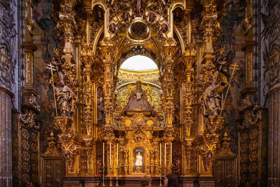 Interior de igreja ornamentado com dourados, um sino grande e estátuas religiosas.