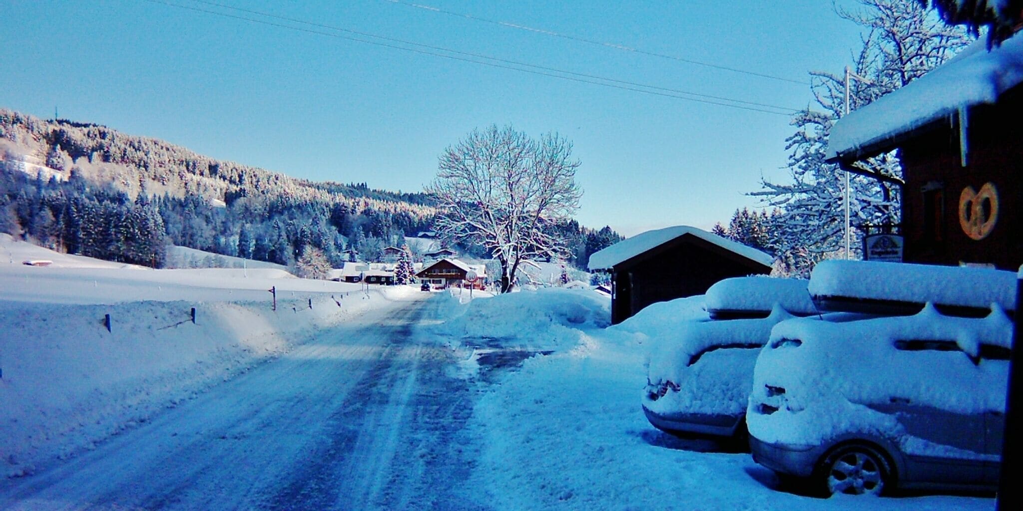 Uma estrada coberta de neve com uma cabana de madeira e carros estacionados em um ambiente rural.