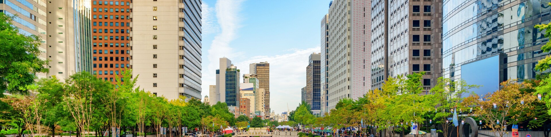 Paisagem urbana com um canal central, edifícios altos e uma passarela para pedestres repleta de vegetação.