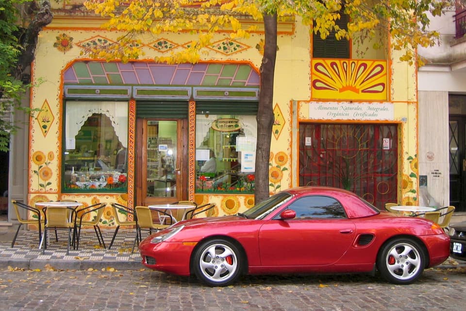 Um carro esportivo vermelho estacionado em frente a um edifício colorido com um desenho de sol na parede e uma placa que diz 'Alimentos Naturales, Integrales, Orgánicos Certificados'.