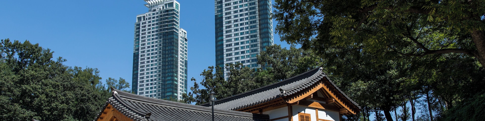 Um edifício tradicional coreano com telhado telhado, rodeado por árvores e arranha-céus modernos ao fundo.