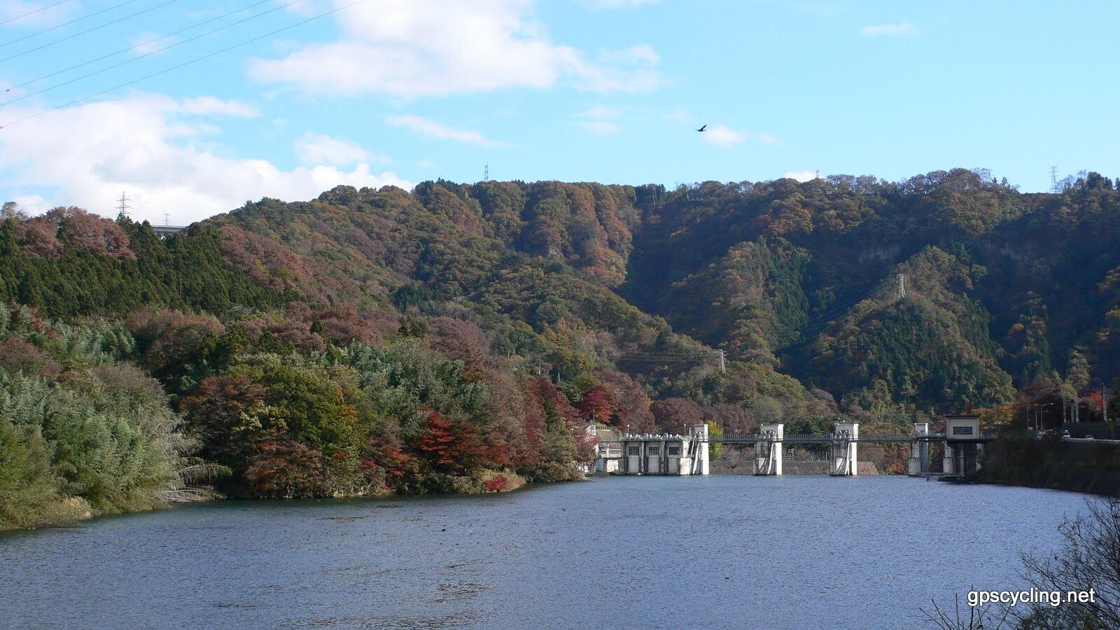 Uma barragem com turbinas de água num rio, rodeado por folhagem de outono e colinas.