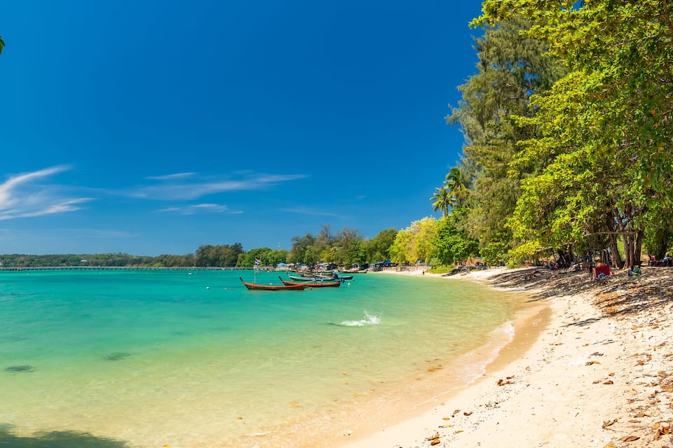 Uma praia turquesa com águas cristalinas, um barco e uma área arborizada.