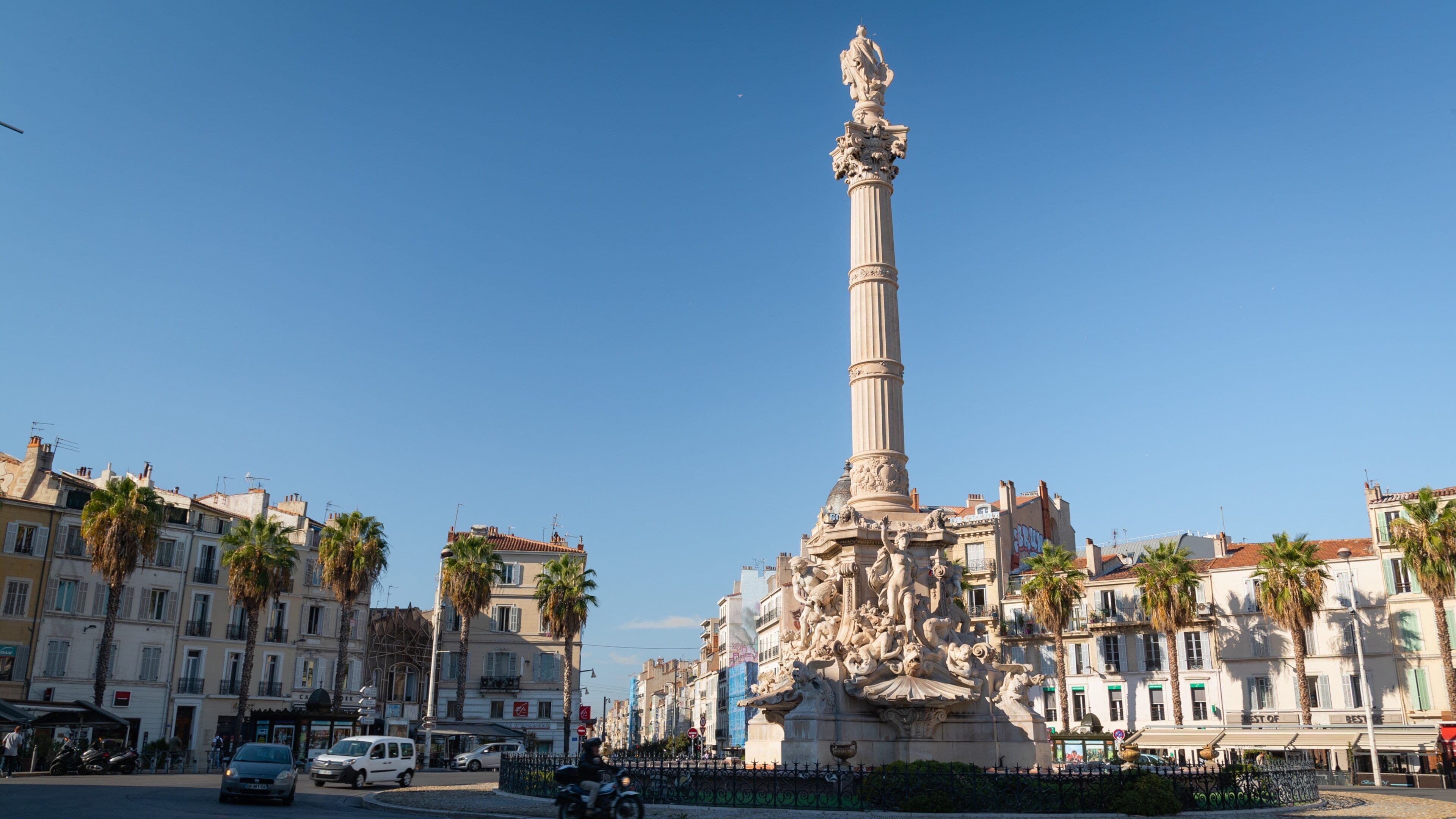 Um monumento alto com esculturas detalhadas, rodeado por palmeiras e edifícios históricos.