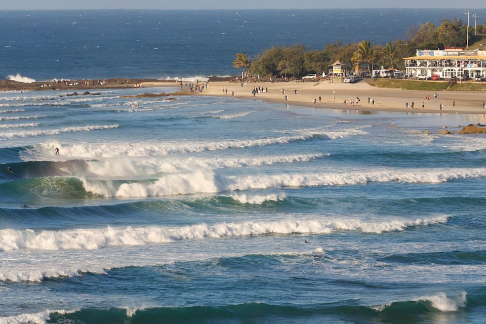 Uma praia costeira com ondas, areal arenoso e um edifício com um letreiro.