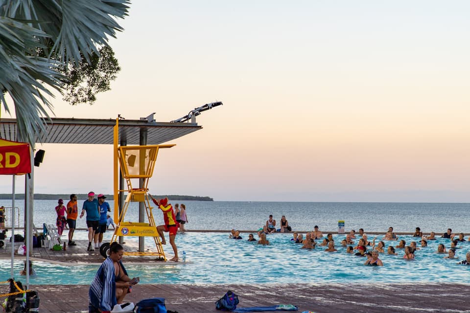 Piscina cheia de gente aproveitando a água, uma cadeira de salva-vidas e um posto de salva-vidas.