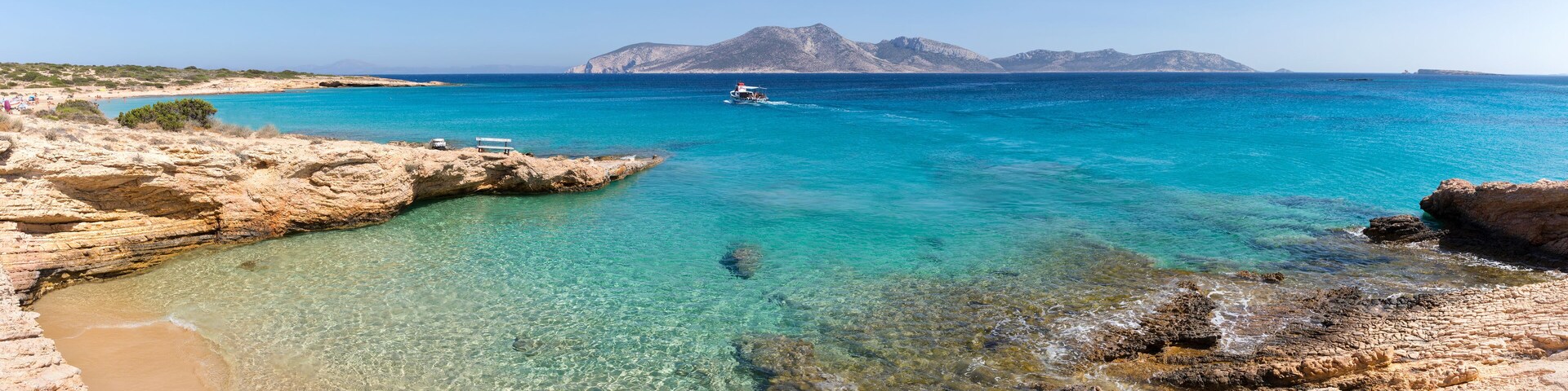Paisagem costeira com águas azul-turquesa cristalinas, formações rochosas e montanhas ao longe.