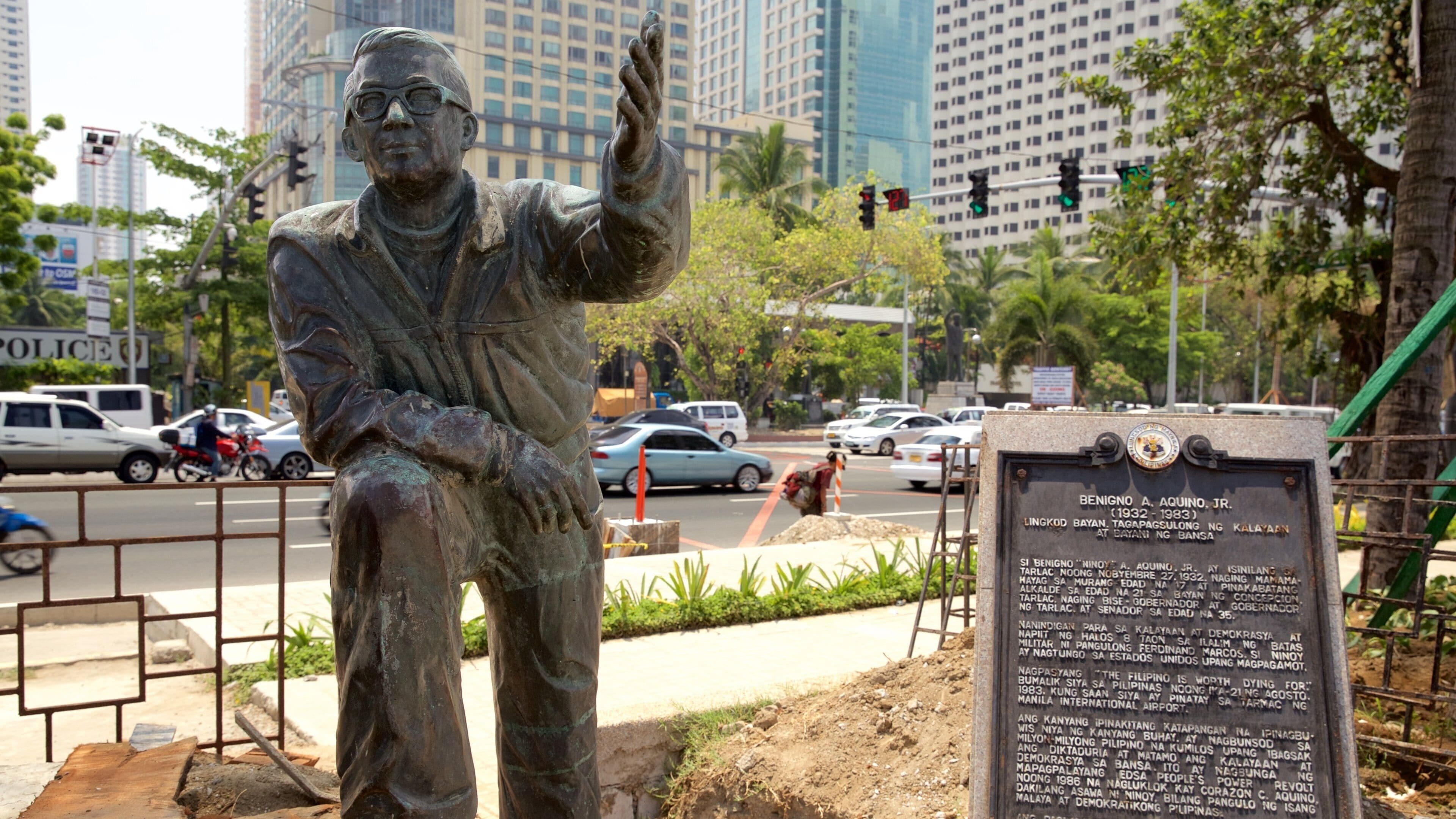 Estátua de um homem com a mão levantada, cercada por uma grade, com uma placa de sinalização ao lado.
