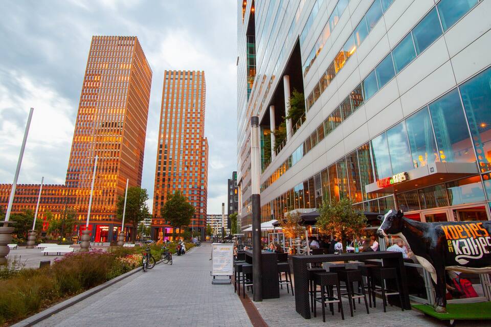 Paisagem urbana com edifícios modernos, mesas ao ar livre num café e uma estátua de vaca.