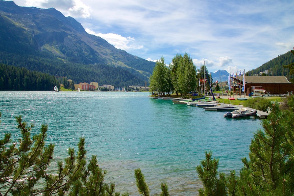 Um lago com barcos, um cais, montanhas e um edifício com bandeiras.