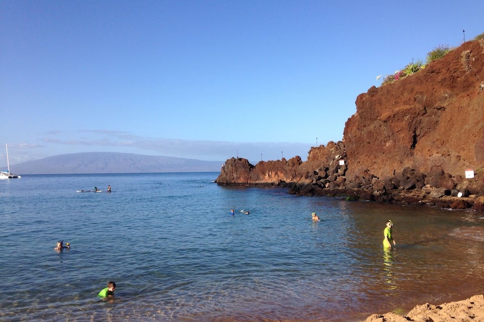 Litoral rochoso com águas azul-claras, um veleiro ao longe e pessoas nadando e entrando no mar.