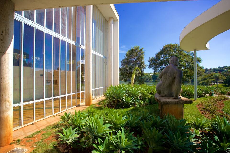 Um edifício moderno com amplas janelas de vidro, um jardim com diversas plantas e uma estátua de pedra.