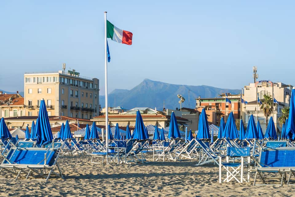 Uma praia com guarda-sóis azuis e espreguiçadeiras, uma bandeira e edifícios ao fundo.