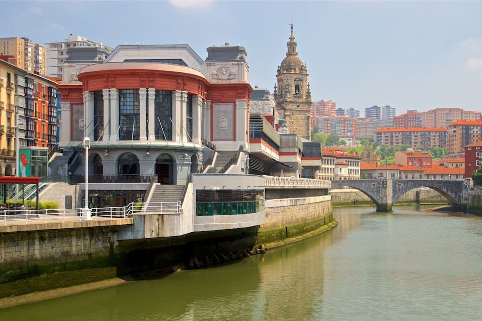 Um rio com uma ponte, um edifício histórico com torre de relógio e arquitetura moderna na margem do rio.