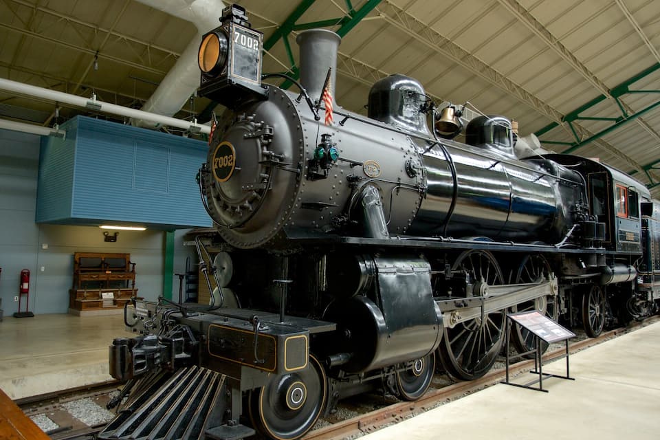 Uma locomotiva a vapor grande e preta com o número 7002, exibida em ambiente interno com teto de metal e paredes azuis.