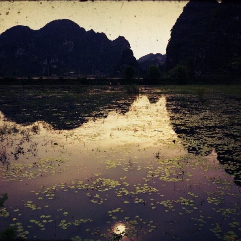 Um rio calmo com folhas de nenúfar e o reflexo das montanhas ao redor.