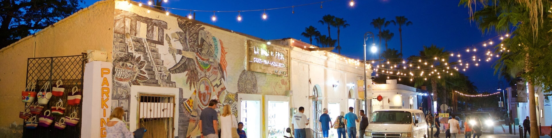 Rua à noite com luzes de Natal, um mural, uma motocicleta e pessoas caminhando.