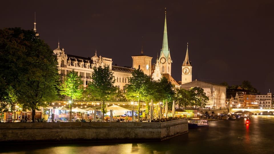 Um centro histórico iluminado à noite, com edifícios destacados, uma torre de igreja imponente e um rio.