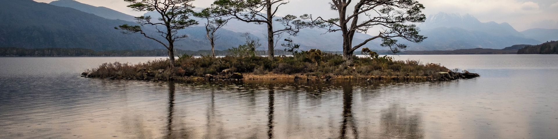 Um lago tranquilo com três árvores numa pequena ilha, rodeado por montanhas.