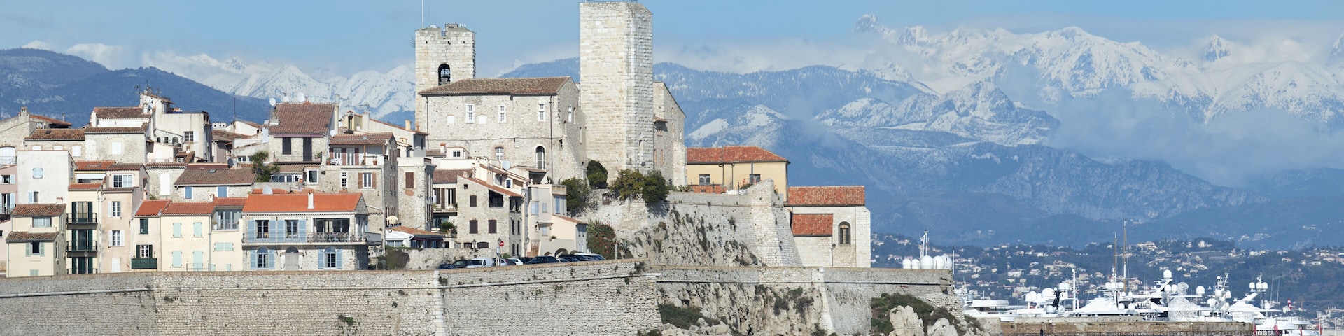 Uma cidade costeira com edifícios históricos de pedra, uma torre imponente e montanhas nevadas ao fundo.