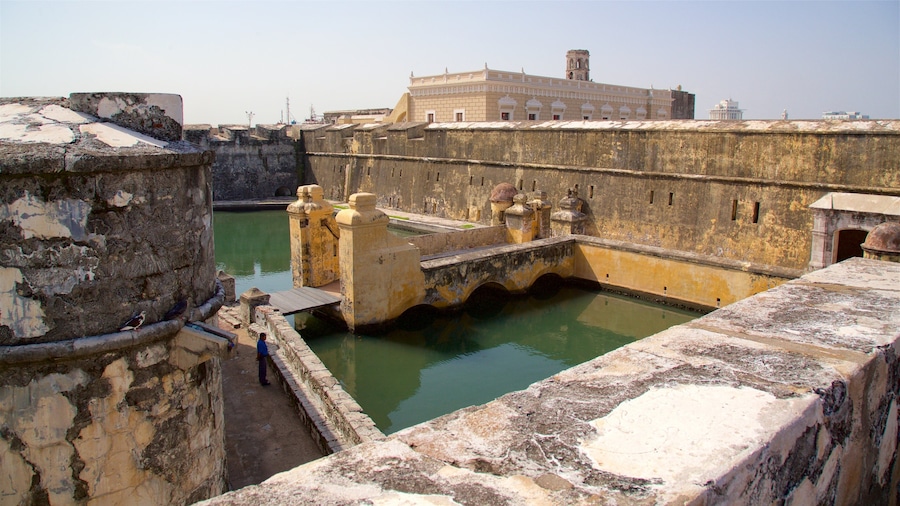 Uma fortaleza histórica com uma ponte sobre um fosso, rodeada por antigas muralhas de pedra e um edifício grande ao fundo.